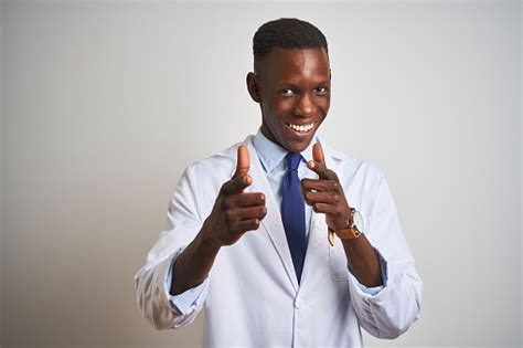 행복하고 재미있는 얼굴로 카메라에 손가락을 가리키는 고립 된 흰색 배경 위에 서있는 코트를 입은 젊은 아프리카 계 미국인 의사 남자 좋은 에너지와 분위기 가리키기에 대한