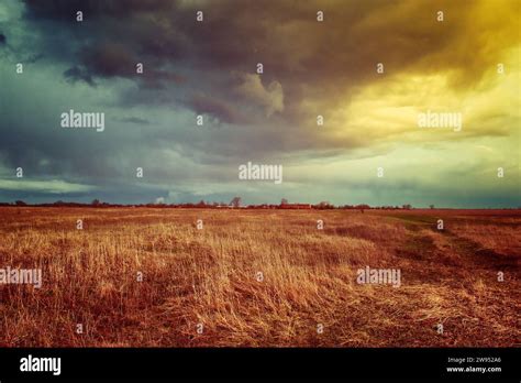 The Image Shows A Vast Field With Dry Grass Under A Dramatic Sky Filled