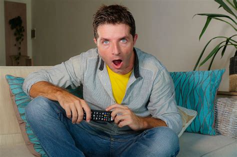 매력적인 영 놀라게 하 고 불신에 깜짝 Tv 뉴스 또는 원격 제어를 들고 거실 소파 소파에서 영화를 보고 연 입으로 캐주얼 옷 집에서 남자 충격 감정에 대한 스톡 사진 및