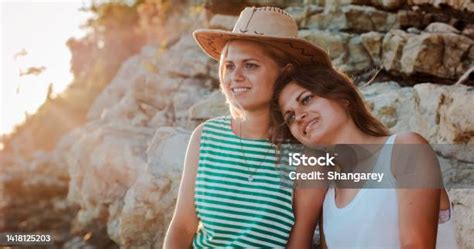 힙스터를 입은 두 명의 젊은 쾌활한 여성이 바다 해안의 바위 위에 모자를 씁니다 소녀 바다 섬 및 오렌지 햇빛이있는 여름 풍경