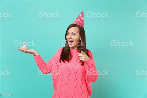 핑크 니트 스웨터에 밝은 젊은 여성 생일 모자 포인팅 검지 카메라에 잡아 손에 케이크 촛불 파란색 벽 배경에 고립 사람들의 라이프 스타일 개념입니다 복사 공간을 비웃는 다