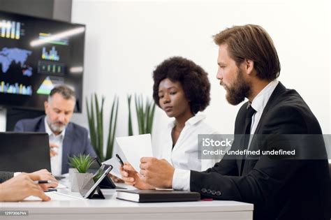 정장 정장 정장에 잘생긴 미소 수염 사업가 그의 남성과 여성 다민족 동료와 테이블에 앉아 통계 차트와 회사의 재무 보고서를 분석 교육에 대한 스톡 사진 및 기타 이미지