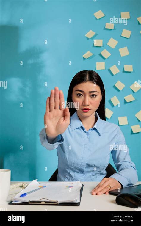 Asian Woman Raising Hand In Refusal Gesture Towards Camera Sitting At