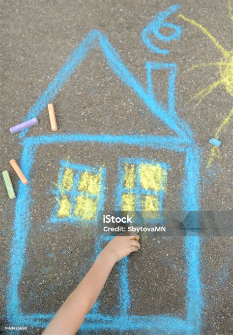 어린 소년은 보도의 아스팔트에 색칠 된 분필로 그려진 집과 태양을 그리고 있습니다 도로의 회색 배경에 있는 아이 창조적인 그림