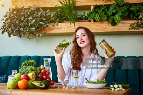 배경에 녹색 꽃과 신선한 과일과 야채와 아름다운 인테리어에 스무디와 신선한 물을 마시는 젊은 여성 건강한 식생활 개념 비건 식사 및 해독 메뉴 가정 생활에 대한 스톡 사진