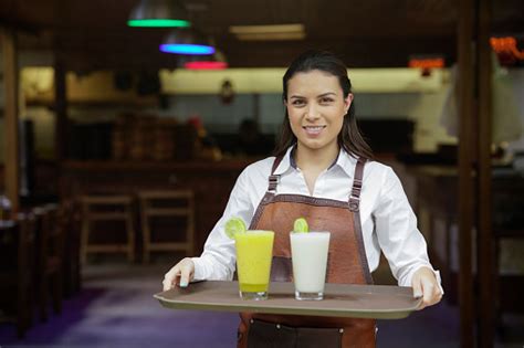 검은 머리의 라틴 여성은 흰색 셔츠와 석영 앞치마를 입고 주문한 가족을 위해 주스 두 잔을 들고 있는 레스토랑 유니폼을 입었습니다 30 39세에 대한 스톡 사진 및 기타