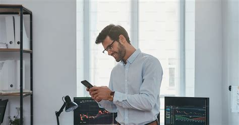 현대 비즈니스 정장을 입은 행복하고 성공적인 트레이더는 투자 목적으로 스마트폰으로 거래 그래프와 보고서를 보고 사무실에 서서 미소를 짓고 있습니다 외환 시장 무역