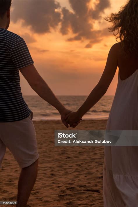 뒷모습 젊은 사랑스러운 가족 커플이 손을 잡고 캐주얼 한 옷을 입은 남자와 여자가 바다 일몰 배경에서 모래 사장을 함께 걷고