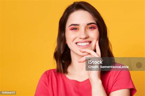 캐주얼 핑크 T 셔츠에 아름 다운 젊은 여자를 웃 고 턱에 손으로 포즈 뺨에 손가락을 유지 좋은 분위기를가지고 행복 외모 즐거운