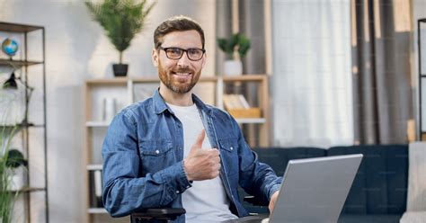 웃는 수염을 기른 남자가 휠체어에 앉아 무릎에 무선 노트북을 들고 있는 동안 엄지손가락을 치켜들고 있다 행복한 장애인 프리랜서가 집에서 일하고 있습니다 사진