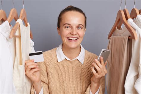 베이지색 스웨터를 입고 회색 벽에 서서 옷을 입은 아름다운 젊은 성인 여성이 선반에 옷장에 걸려 있고 휴대폰과 신용 카드를 들고 이빨 미소로 카메라를 바라보고 있다 30