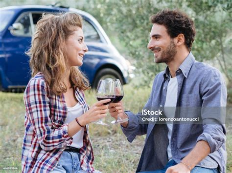 이탈리아 토스카나 시골에서 피크닉을 즐기며 유리 잔에서 레드 와인을 마시는 젊은 연인 부부 늦은 여름 날 올리브 나무 들 사이 잔디에 앉아 그들 뒤에 오래 된 푸른 자동차