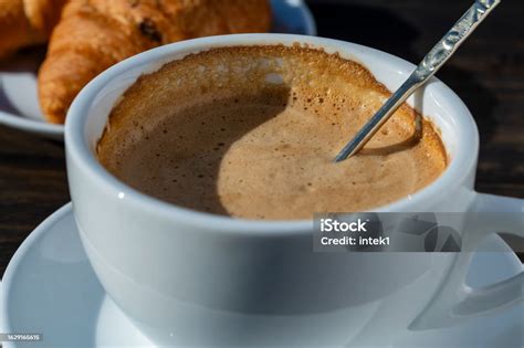 뜨거운 커피 카푸치노의 두꺼운 거품과 하얀 컵에 숟가락과 나무 테이블에 있는 크루아상 클로즈업 우유 거품 카푸치노 라떼 갈색에