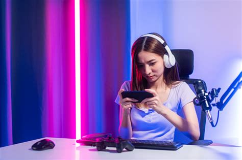휴대 전화에서 비디오 게임 승리 거실에서 스마트 폰을 들고 의자에 앉아있는 젊은 아시아 예쁜 여자 행복한 여성 프로 스트리머 중국어 착용 헤드폰 네온 라이트와 온라인 게임을