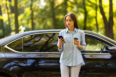 휴대 전화와 커피 컵을 가진 젊은 여자는 전기 자동차 충전 근처에 서 있습니다 태양광 발전소 근처의 공공 충전소에서 차량 충전 친환경 대체 에너지 운송 금속에 대한 스톡