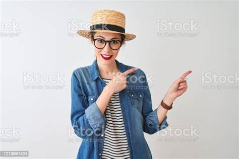 데님 셔츠 안경과 모자를 쓰고 고립된 흰색 배경위에 미소를 지으며 두 손과 손가락으로 측면을 가리키는 카메라를 바라보는 빨간