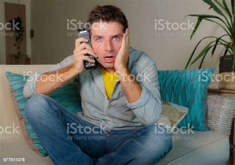 매력적인 영 놀라게 하 고 불신에 깜짝 Tv 뉴스 또는 원격 제어를 들고 거실 소파 소파에서 영화를 보고 연 입으로 캐주얼 옷 집에서 남자 충격 감정에 대한 스톡 사진 및