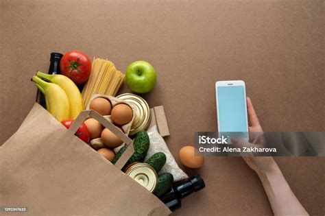음식용품이 담긴 종이 봉투와 중성 베이지색 배경에 전화로 여성손 음식 배달 검역 기부 코로나 바이러스 개념에 대한 모바일 주문 응용 프로그램 텍스트 모형을 위한 공간 복사