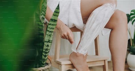 흰 수건을 쓴 젊은 여자가 녹색 식물이 있는 집 욕실에서 다리에 면도 크림을 바르고 있다 피부 관리 및 웰빙 개념 크림으로 피부에 플라스틱 면도기를 들고 손 사진