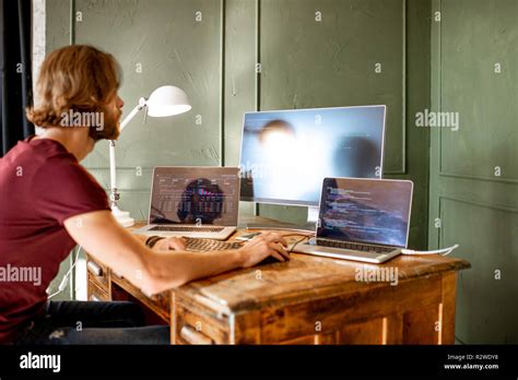 Young Programmer Writing A Program Code Sitting At The Workplace With Three Monitors In The