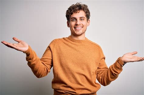 젊은 금발의 잘생긴 남자 와 곱슬 머리 캐주얼 스웨터 위에 흰색 배경 미소 보여주는 양손 오픈 손바닥 제시 및 광고 비교 및 균형 결정에 대한 스톡 사진 및 기타 이미지