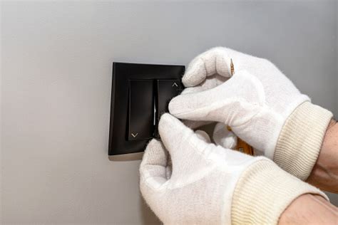 전기 기술자가 롤러 셔터 스위치의 검은 색 버튼을 창문 옆 방에 흰색 장갑으로 위아래로 장착합니다 남자에 대한 스톡 사진 및