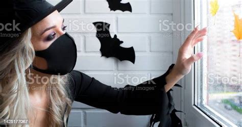 검은 마녀 의상을 입은 슬픈 금발과 얼굴 마스크가 창문 밖을 바라 봅니다 코비드로부터 할로윈 보호 준비 10월에 대한 스톡 사진 및 기타 이미지 Istock