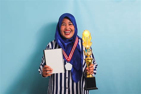 히잡을 쓴 중년의 아시아 사업가가 책과 금 트로피를 들고 있고 파란색 배경에 고립되어 있다 1위에 대한 스톡 사진 및 기타 이미지 Istock