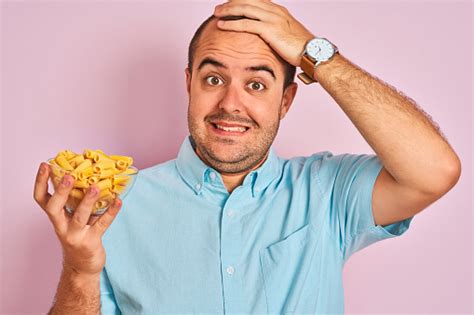 마카로니 파스타와 그릇을 들고 젊은 남자는 머리에 손으로 스트레스 부끄러움과 깜짝 얼굴 분노와 좌절에 충격 고립 된 분홍색 배경 위에 서 실수에 대한 두려움과 화가 감정에