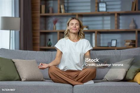 거실의 소파에 앉아 눈을 감고 명상을 하고 있는 집에서 쉬고 있는 노인 여성 연꽃 포즈를 취한 성숙한 금발의 주부 명상하기에