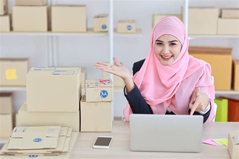 무슬림 정장을 입은 활동적인 아시아 여성이 앉아서 컴퓨터 모바일 스마트폰 온라인 소포 상자 배달을 하고 있다 스타트업 중소기업 프리랜서 일하는 소녀가 손에 무언가를 보여주고