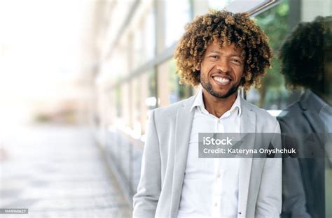 곱슬 머리를 가진 행복한 잘 생긴 자신감있는 긍정적 인 아프리카 계 미국인 또는 브라질 남자 세련된 정장을 입고 야외에 서서 카메라를보고 친절하게 웃고 있습니다 복사 공간