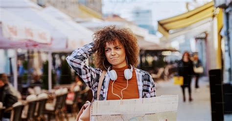 곱슬 머리와 격자 무늬 셔츠를 입고 도시지도를 들고 메인 스트리트에 서있는 동안 어디로 가야할지 찾고있는 젊은 혼혈 여성 관광객을 잃었습니다 사진 Unsplash의