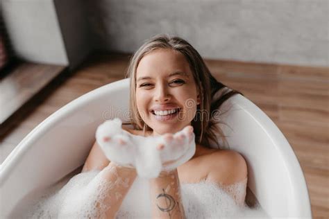 Playful Lady Having Fun While Lying In Bathtub Full Of Foam Blowing Soap Bubbles And Smiling