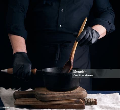 검은 색 유니폼과 라텍스 장갑을 입은 남성 요리사가 손잡이가달린 주철 팬에 빈티지 나무 숟가락 음식을 저어줍니다 가정용 장비에