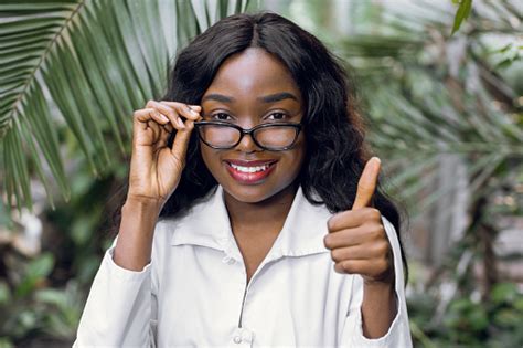 젊은 성공적인 아프리카 여성 연구원 또는 흰색 코트를 입은 농학자 이국적인 열대 식물과 야자수가있는 아름다운 온실에 실내에 서서