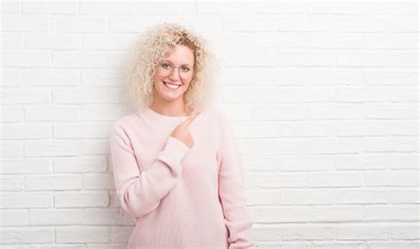 흰색 벽돌에 곱슬 머리 젊은 금발 여자 손과 카메라 보고 얼굴에 행복 하 고 자연 식 가진 측까지 손가락으로 가리키는 얼굴의 미소와 쾌활 한 벽 가리키기에 대한 스톡 사진