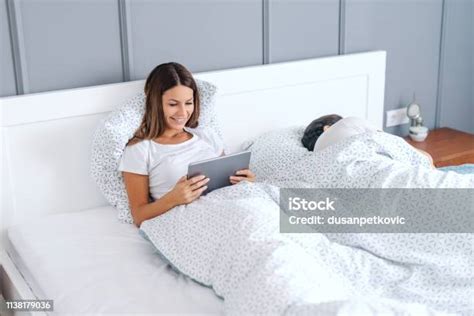 태블릿을 사용 하 여 화려한 백인 갈색 머리와 그의 남편이 그녀의 옆에 잠자는 동안 아침에 침대에 앉아 침실 인테리어입니다 가정 생활에 대한 스톡 사진 및 기타 이미지