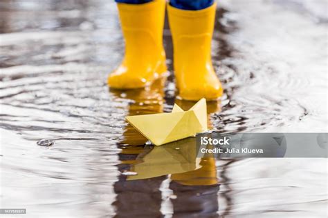 봄입니다 노란 고무 장화를 신은 작은 아이가 웅덩이를 뛰어 넘고 놀고 물 위에서 종이 보트를 발사합니다 봄과 가을 휴가의 사진 봄의 개념 계절에 대한 스톡 사진 및 기타
