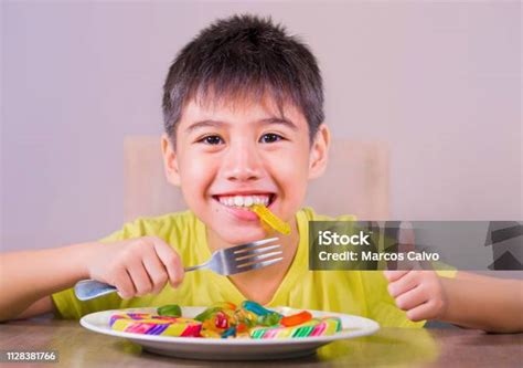 젊은 행복 하 고 흥분 남자 아이 명랑 먹는 접시 가득 사탕과 막대 사탕 설탕 중독 및 남용과 달콤한 건강에 해로운 영양에 회색 배경에 고립 된 테이블에 앉아 웃 고 건강에