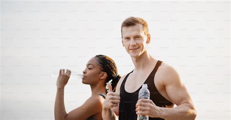운동복을 입은 다민족 커플은 야외에 서서 운동 중에 휴식을 취하고 있습니다 물을 마시는 아프리카 여자 엄지손가락을 치켜든 백인 남자 사진 Unsplash의 건강한