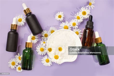 카모마일 꽃이있는 파스텔 보라색 보라색 배경에 물방울이있는 어두운 유리 화장품 병 피부관리를 위한 천연 보습 화장품의 개념