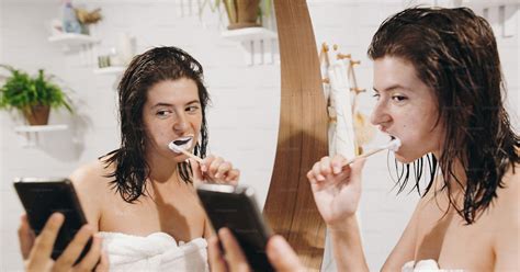 소셜 미디어가 영향을 미칩니다 하얀 수건을 쓴 젊은 행복한 여자가 이를 닦고 둥근 거울에 있는 세련된 욕실에서 전화 화면을 보고 있다 날씬한 섹시한 여자 샤워 후 일상