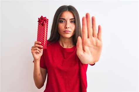 진지하고 자신감있는 표정 방어 제스처와 정지 기호를하고 열린 손으로 고립 된 흰색 배경 위에 서 발렌타인 선물을 들고 젊은 아름다운 여자 갈색 머리에 대한 스톡 사진 및