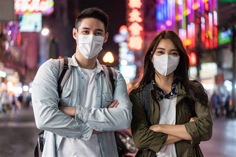 밤에 신혼 여행을 위해 도시에서 여행하는 아시아 커플의 초상화 매력적인 젊은 남자와 여자 배낭 여행자 관광 착용 마스크 함께 휴가에 시간을 보내고 카메라를보고 즐길 수