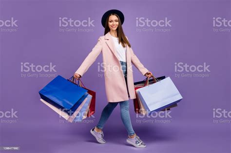 풀 길이 프로필 측면 사진 의 쾌활한 사랑스러운 트렌디 한 소녀 잡고 많은 가방은 보라색 색상 배경에 고립 된 쇼핑몰 착용 베이지 색 의상 데님 모자 운동화에서 이동 갈색