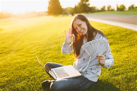 달러 현금 돈을 여성 지주 번들 여자 잔디에 앉아 푸른 잔디 햇빛 잔디 야외에 도시 공원에서 노트북 Pc 컴퓨터에서 작업 모바일 사무실입니다 프리랜서 비즈니스 개념입니다