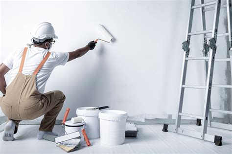 한 남자가 롤러로 하얀 벽을 그립니다 내부의 수리 빈 방에 벽을 그리는 젊은 남성 데코레이터 개념 빌더 또는 빈 방에 페인트 롤러와 헬멧에 화가 Diy에 대한 스톡 사진 및