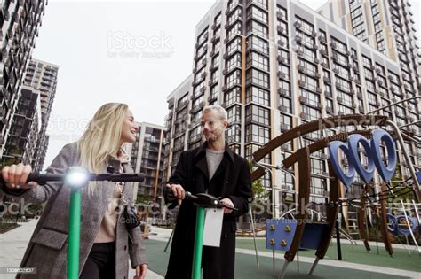 젊은 부부는 전기 스쿠터에 좋은 시간을 보내고 배경에 현대 아파트 블록 남자와 여자는 전자 스쿠터를 임대 빠른 여행 개념 친환경 교통 대도시 개념의 Autmn 20 24세에