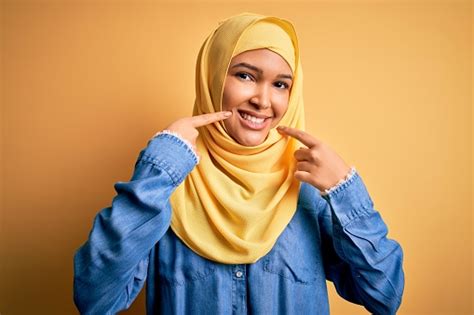 노란 배경에 아랍 전통 히잡을 입고 곱슬 머리를 가진 젊은 아름다운 여성은 손가락 이빨과 입으로 가리키는 쾌활한 모습과 미소를 짓고 있습니다 치과 건강 개념 가리키기에 대한